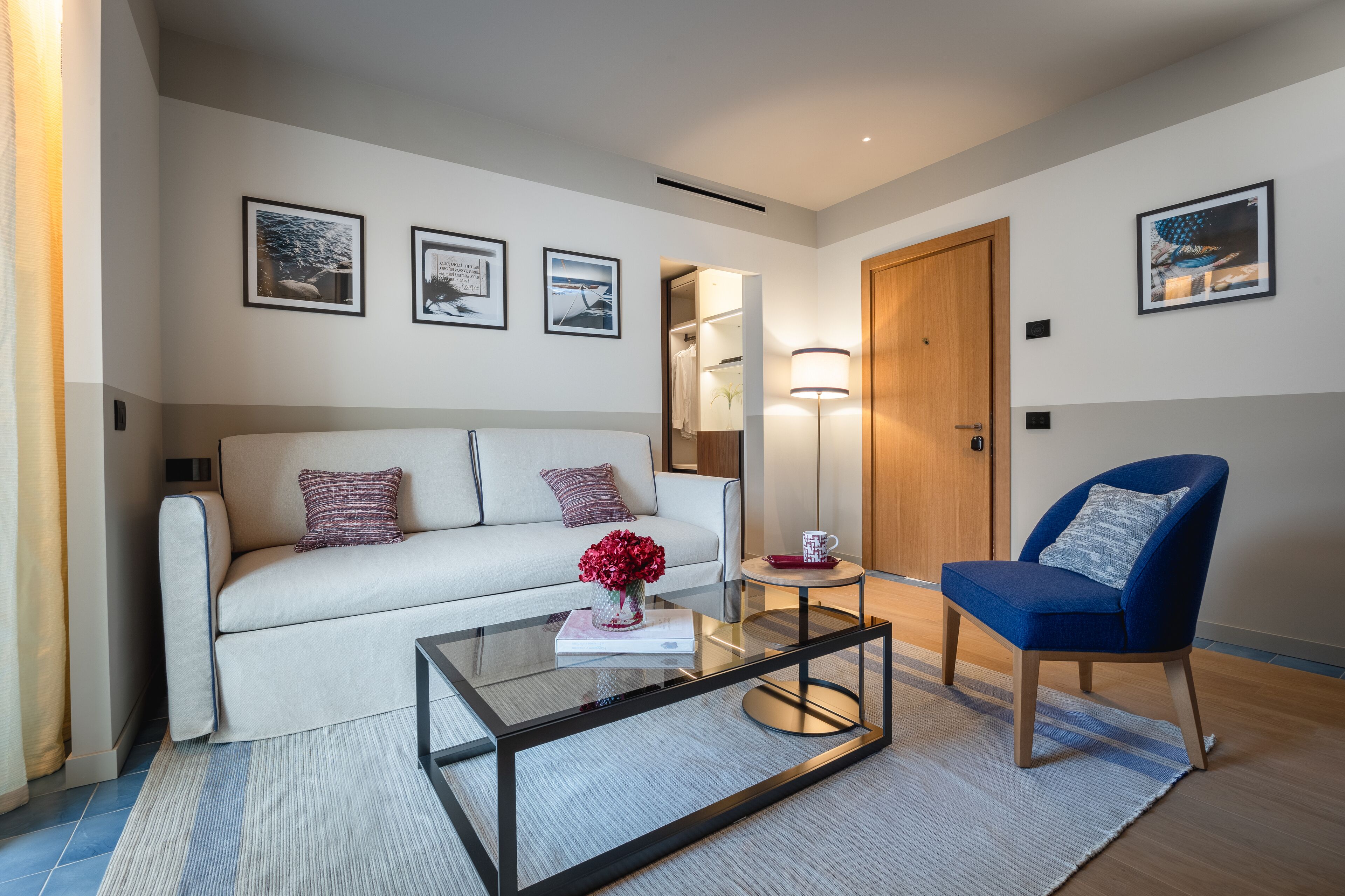 Uma sala de estar moderna com um sofá, uma mesa de centro de vidro, uma poltrona azul e uma pequena mesa lateral.