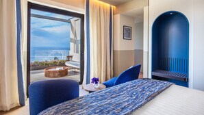 Quarto de hotel moderno com uma cama, uma poltrona azul, uma mesinha com um vaso de flores e vista para a cidade através de grandes janelas.