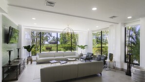 Uma sala de estar espaçosa com um sofá grande, uma mesa de centro e uma TV fixada na parede. Há janelas amplas com vista para o exterior.