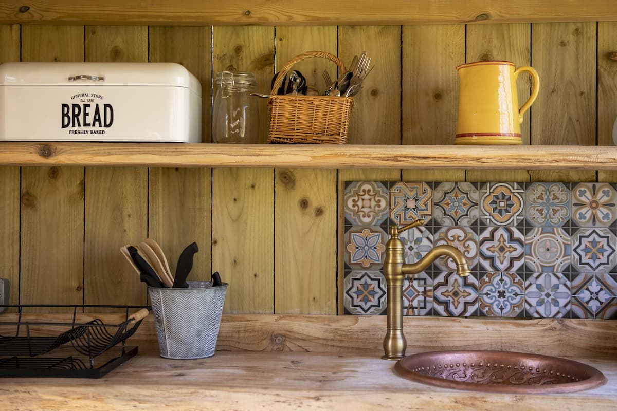 Uma cozinha com um aparador de pão, uma cesta de vime com utensílios, uma caneca amarela, azulejos estampados no painel e uma pia de cobre.