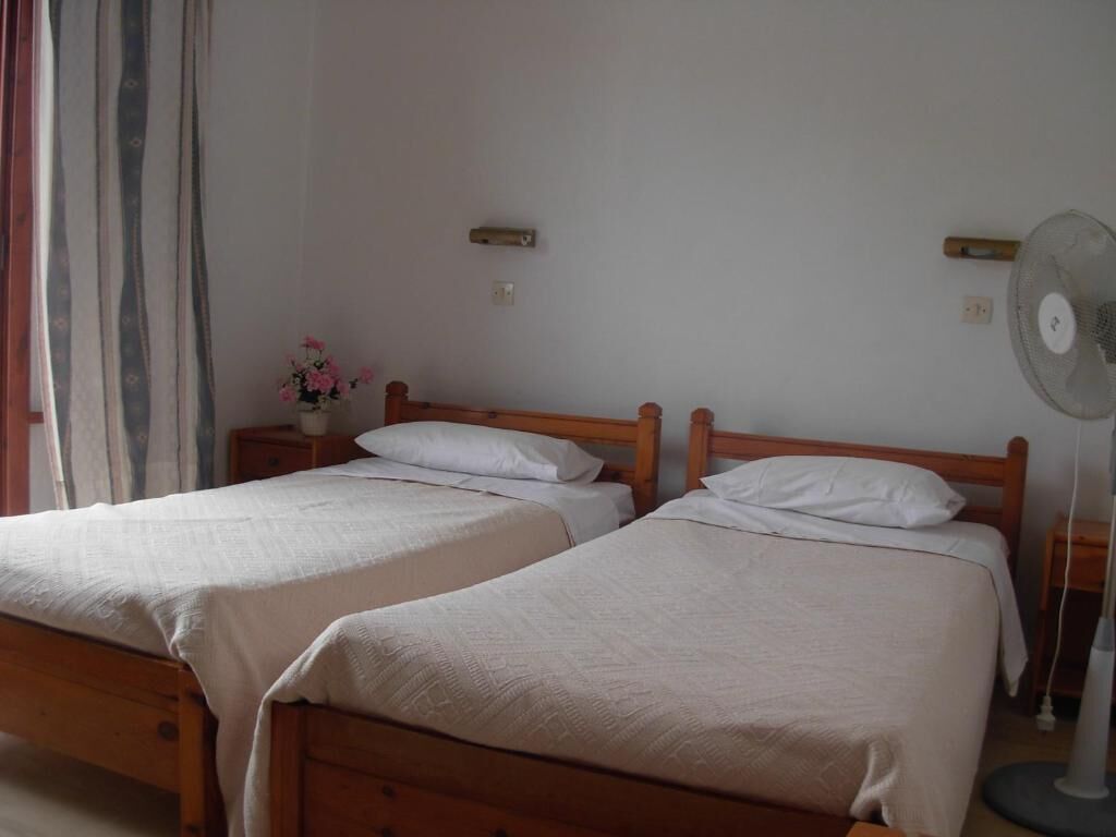 Quarto com duas camas de madeira, uma pequena mesa de cabeceira com um vaso de flores, um ventilador fixado na parede e uma janela com cortinas.