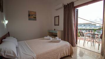 Quarto de hotel com cama, mesa de cabeceira, pintura, janela com cortinas e varanda com cadeiras e guarda-sóis.