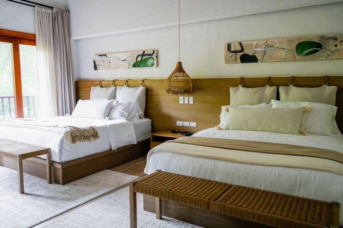 Quarto de hotel com duas camas, cabeceira de madeira, um banquinho de fibra natural e arte abstrata nas paredes.