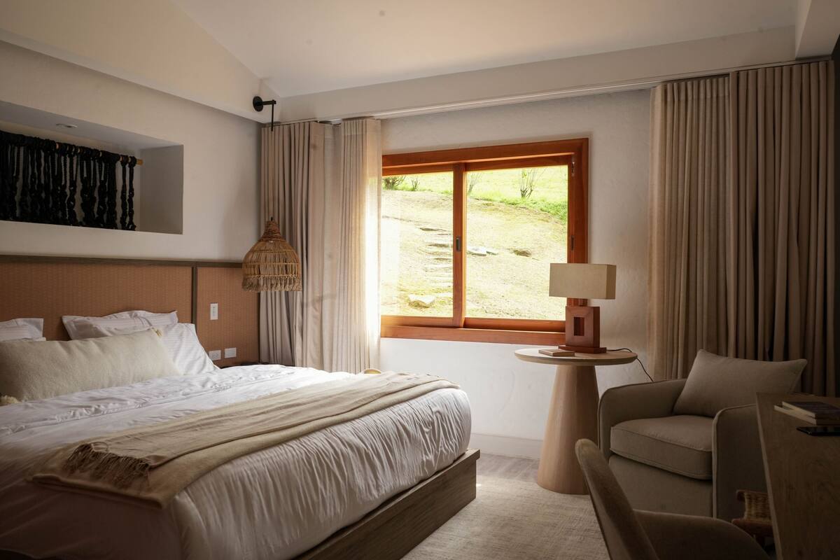 Quarto de hotel com uma cama grande, uma mesinha redonda com abajur, um sofá e uma janela com cortinas.