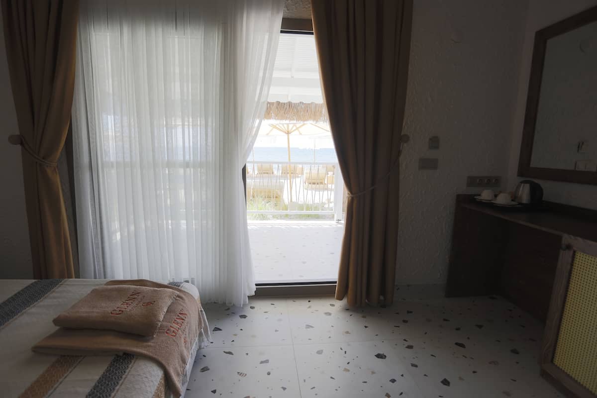 Quarto de hotel com cama, uma pequena escrivaninha, espelho e vista para o mar através de uma janela com cortinas.