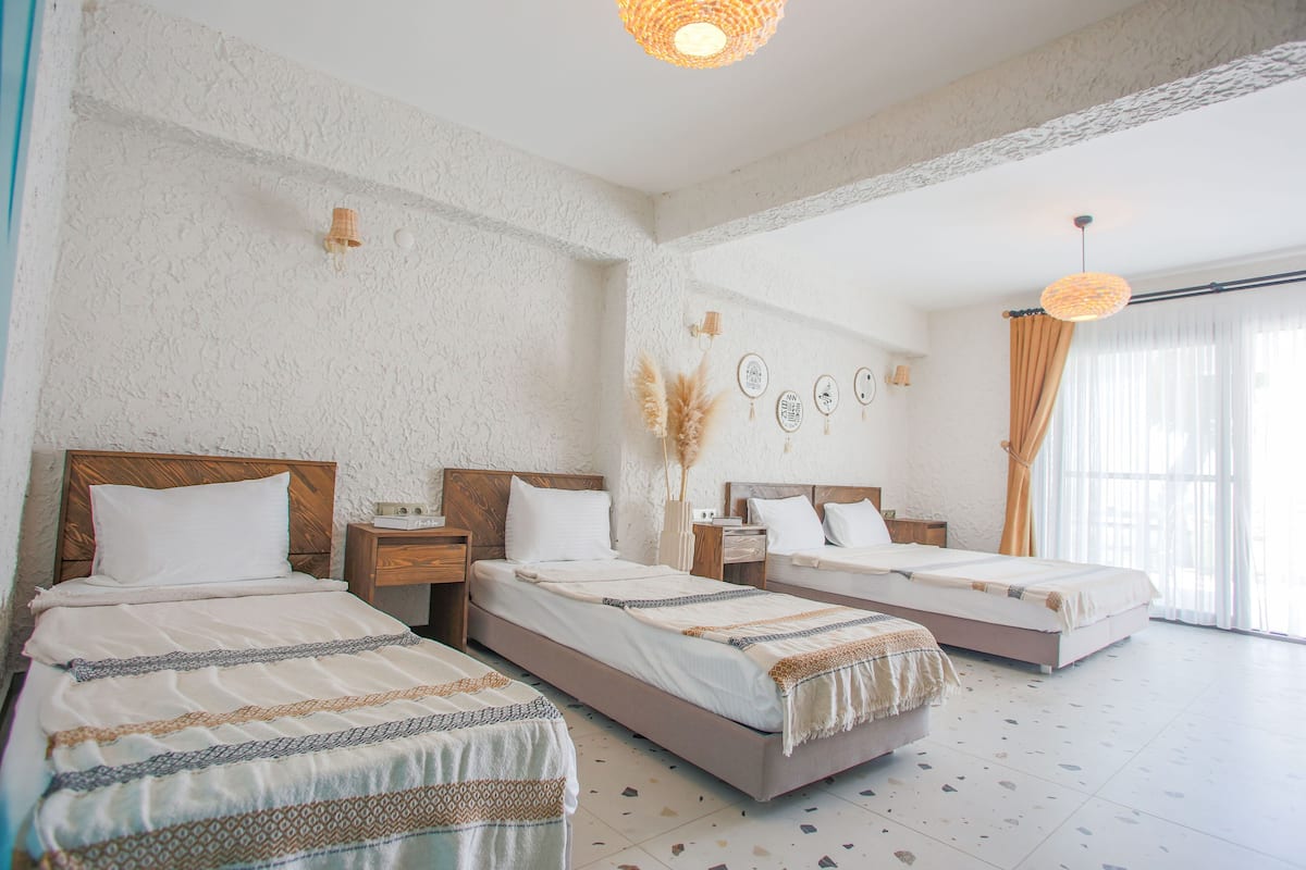 Quarto de hotel com três camas, uma janela grande com cortinas e parede com textura.