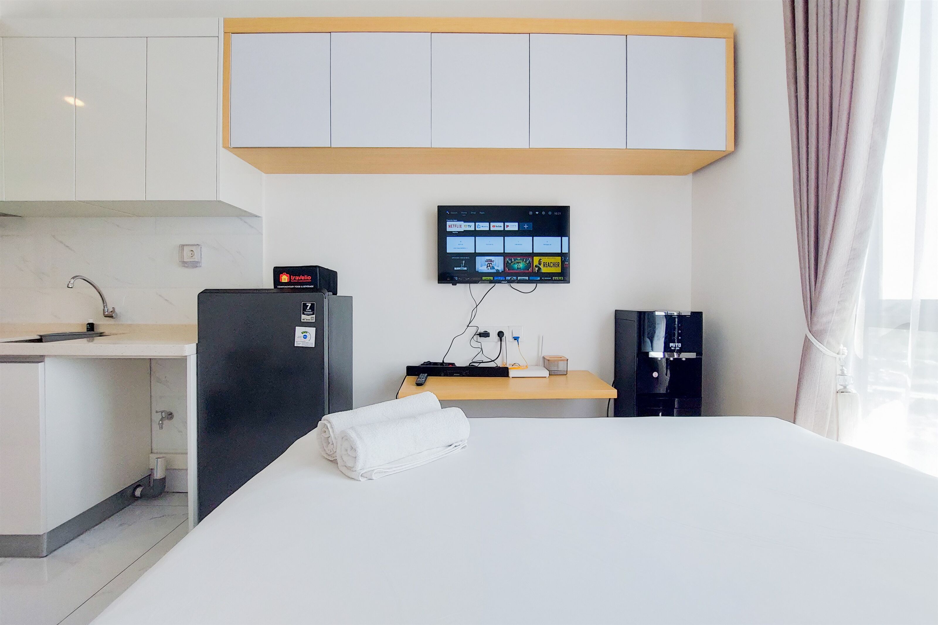 Quarto de hotel com cama, escrivaninha, televisão, minibar e janela com cortinas.