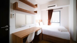 Quarto de hotel com escrivaninha de madeira, cama com roupa de cama branca, um espelho grande e um ar condicionado.