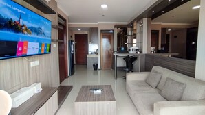Uma sala de estar moderna com uma TV de tela plana, um sofá, uma mesa de centro e uma cozinha ao fundo.