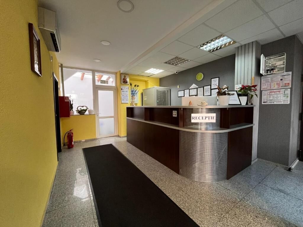 Uma área de recepção com balcão identificado como "RECEPTION", um extintor de incêndio e um tapete preto que leva à entrada.