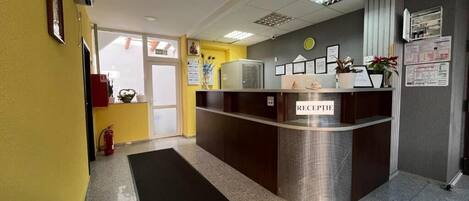 Uma área de recepção com balcão identificado como "RECEPTION", um extintor de incêndio e um tapete preto que leva à entrada.