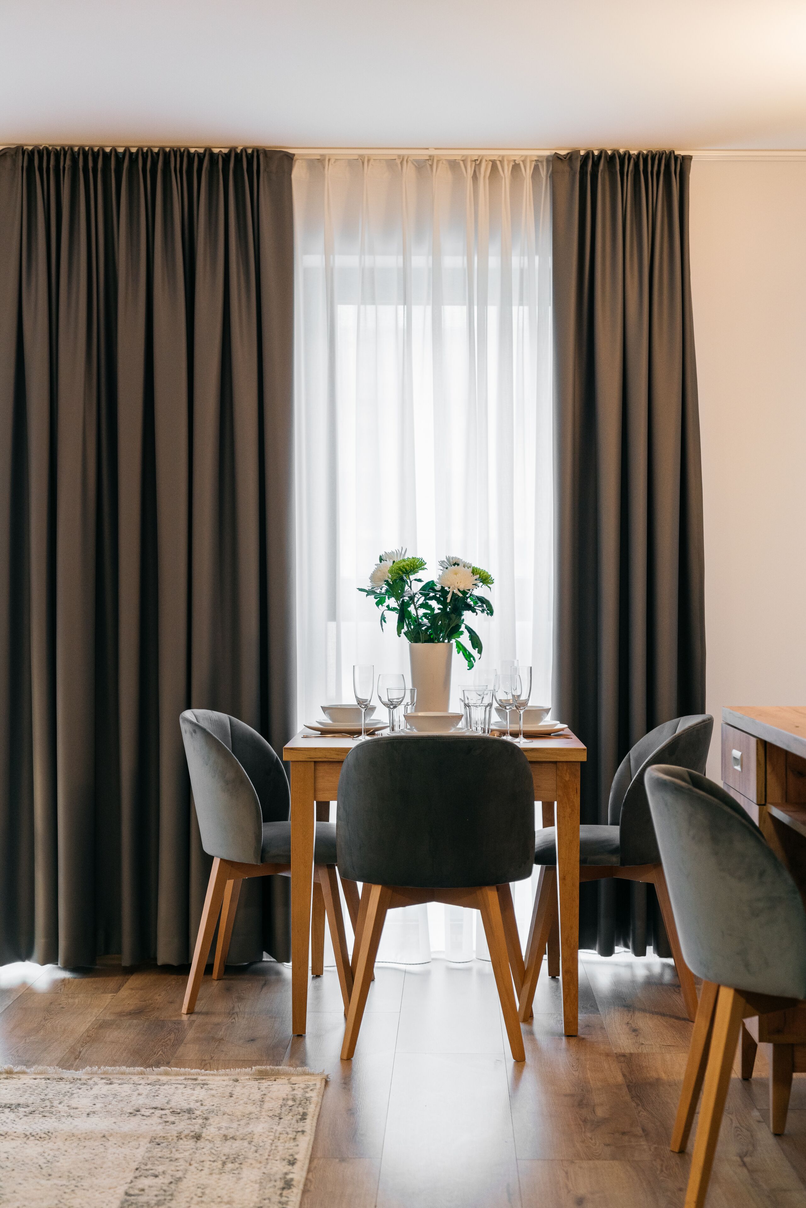 Uma área de refeições com uma mesa posta para dois, uma planta em vaso e uma janela com cortinas.