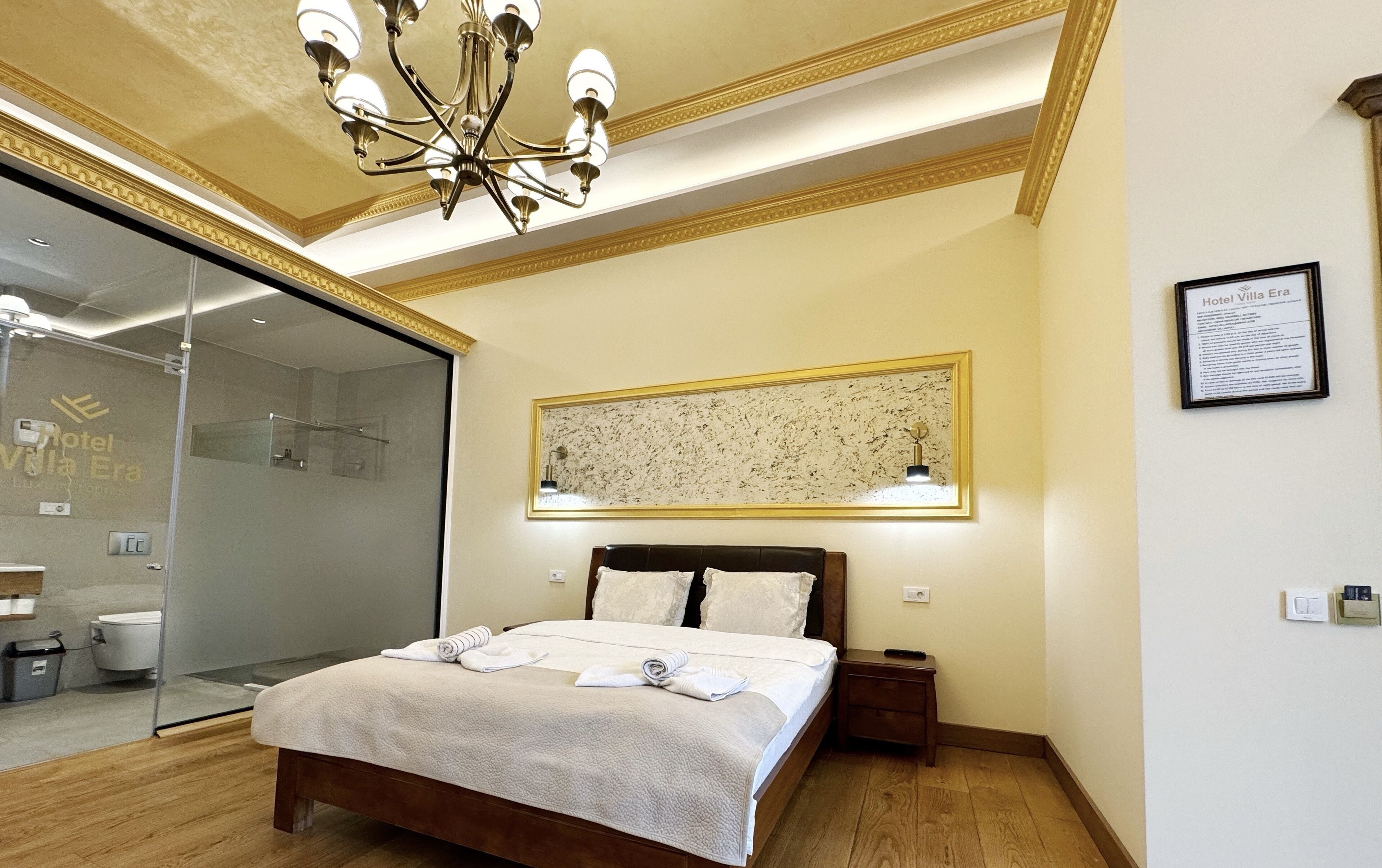 Quarto de hotel com uma cama grande, um candelabro, box de chuveiro de vidro e uma obra de arte emoldurada na parede.