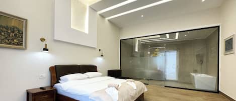 Quarto de hotel moderno com uma cama grande, mesas de cabeceira, um criado-mudo, uma luminária na parede e uma janela ampla.