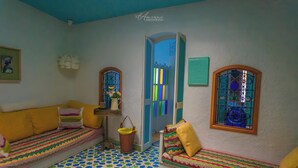 Quarto com piso estampado, um sofá com almofadas, uma mesinha de centro com um vaso e uma janela com grades de ferro decorativas.