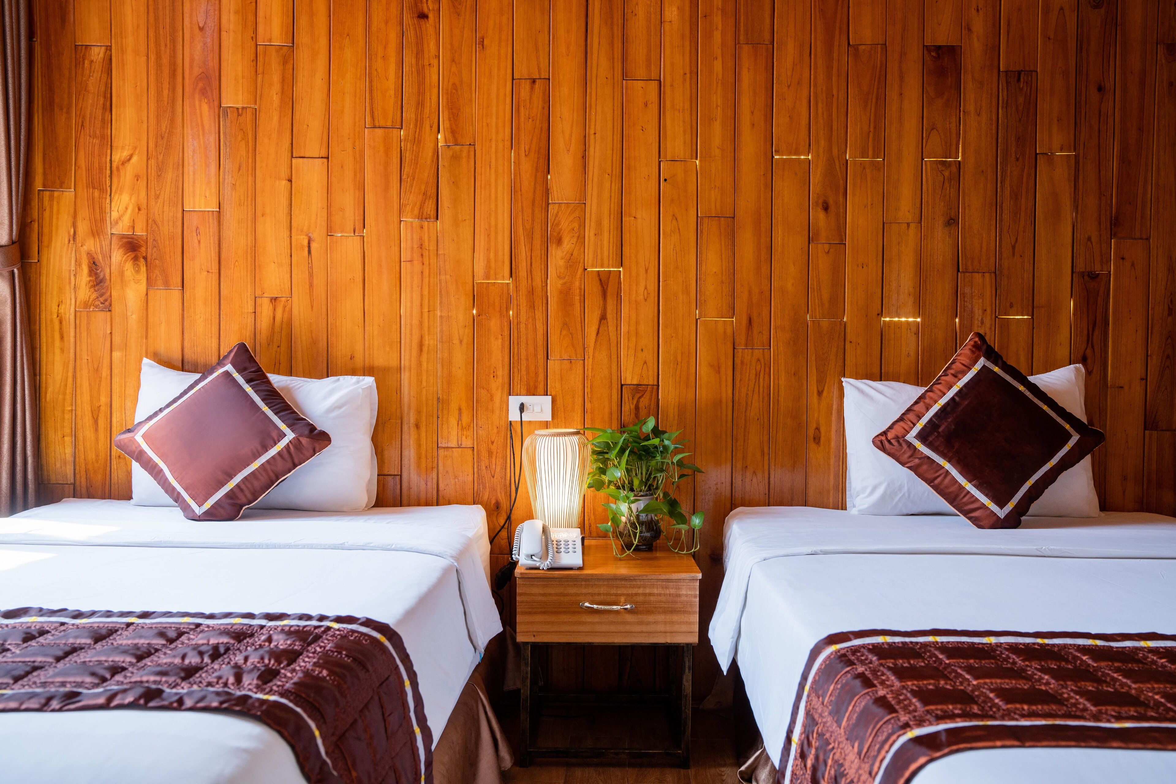 Quarto de hotel com duas camas, paredes de painel de madeira, mesa de cabeceira com abajur, telefone e uma planta.