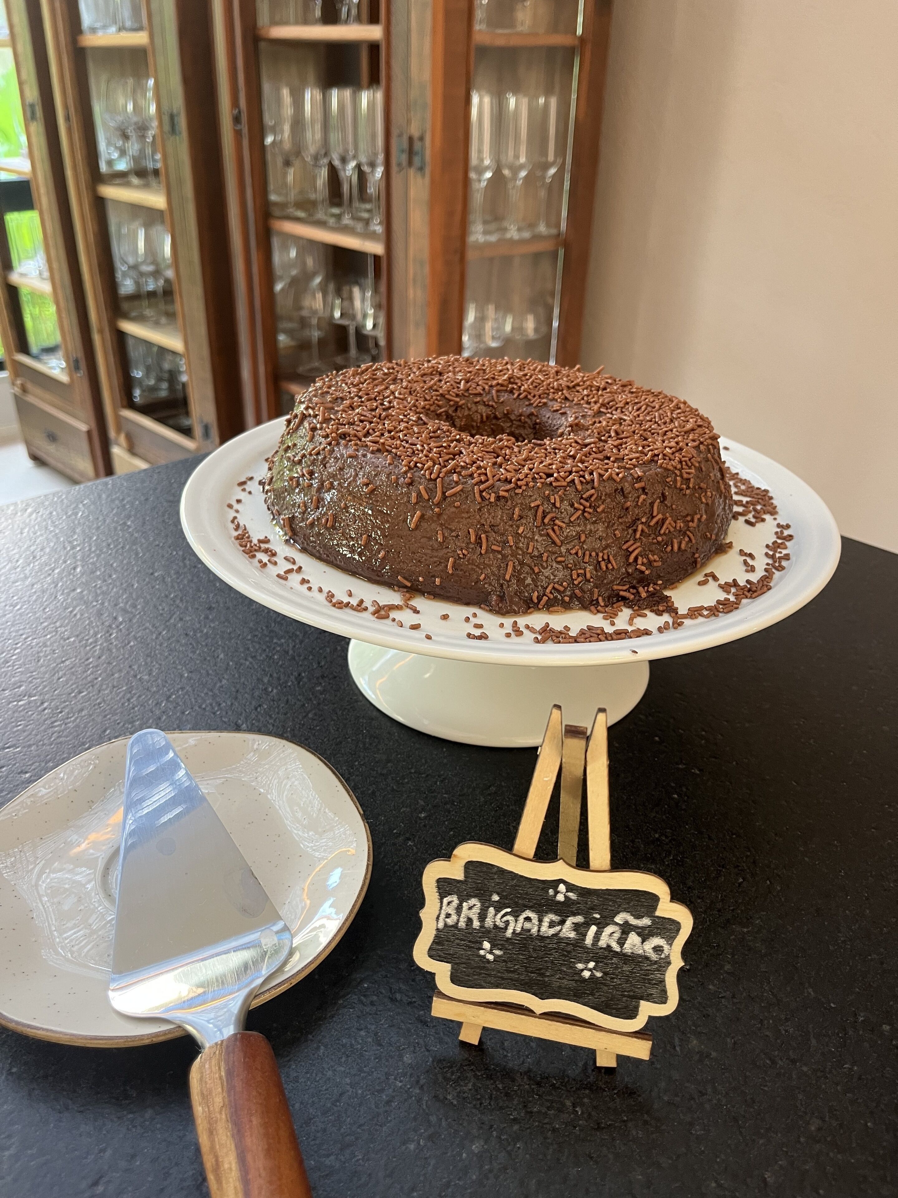 Um bolo bundt de chocolate sobre um prato branco, com raspas de chocolate, um garfo para bolo e uma placa com a inscrição 'BRIGADE 1'.