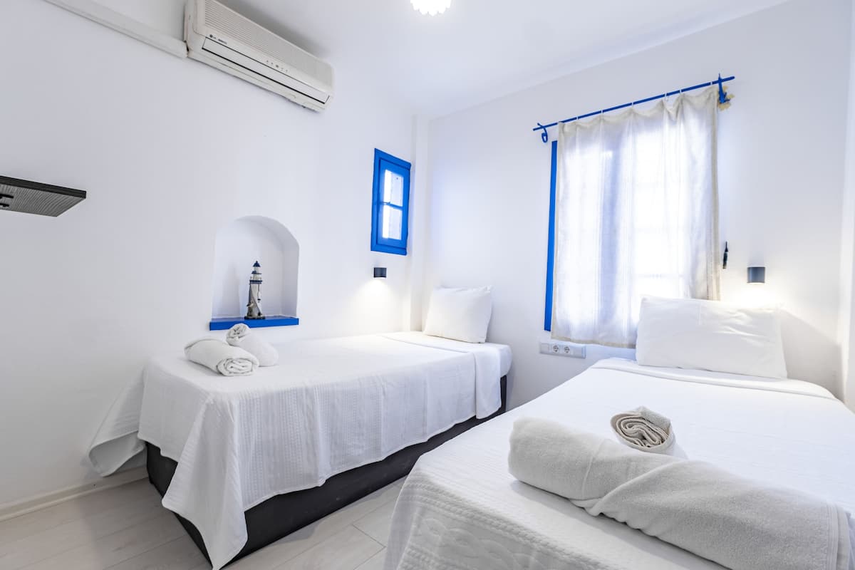 Quarto de hotel com duas camas, uma janela com cortinas azuis, uma mesinha com luminária e um ar condicionado fixado na parede.