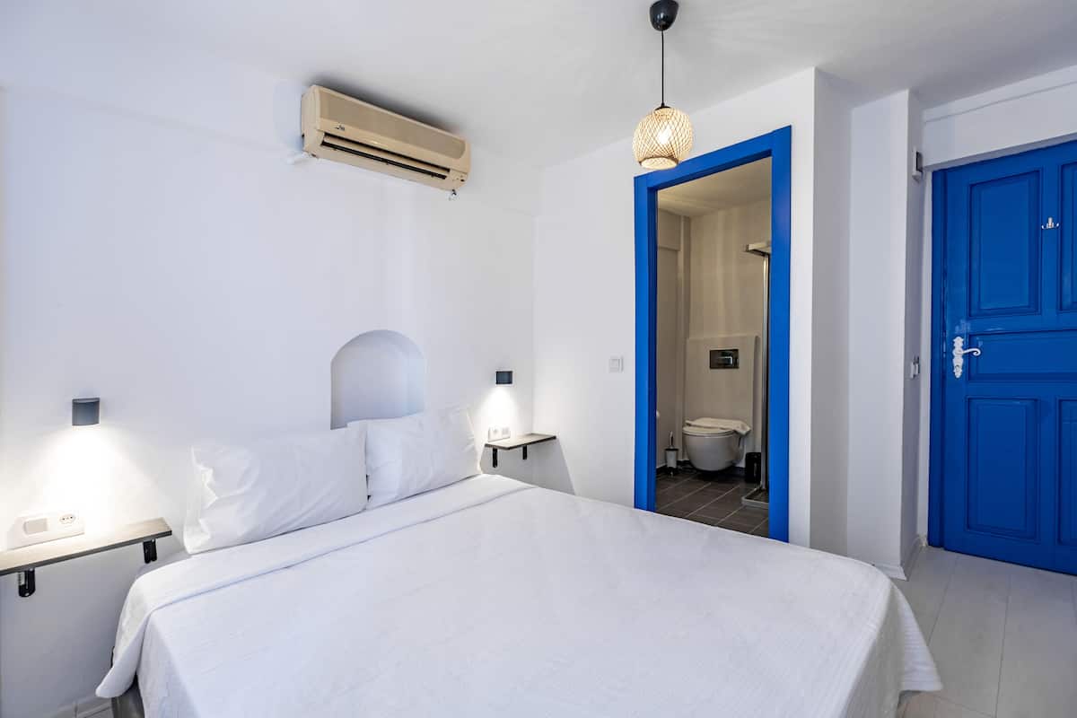 Quarto de hotel com uma cama, mesas de cabeceira, aparelho de ar condicionado, banheiro visível através de uma porta aberta e uma porta azul.