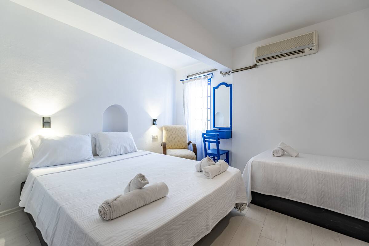 Quarto de hotel com uma cama grande, uma cama menor, uma cadeira, uma escrivaninha e um espelho.