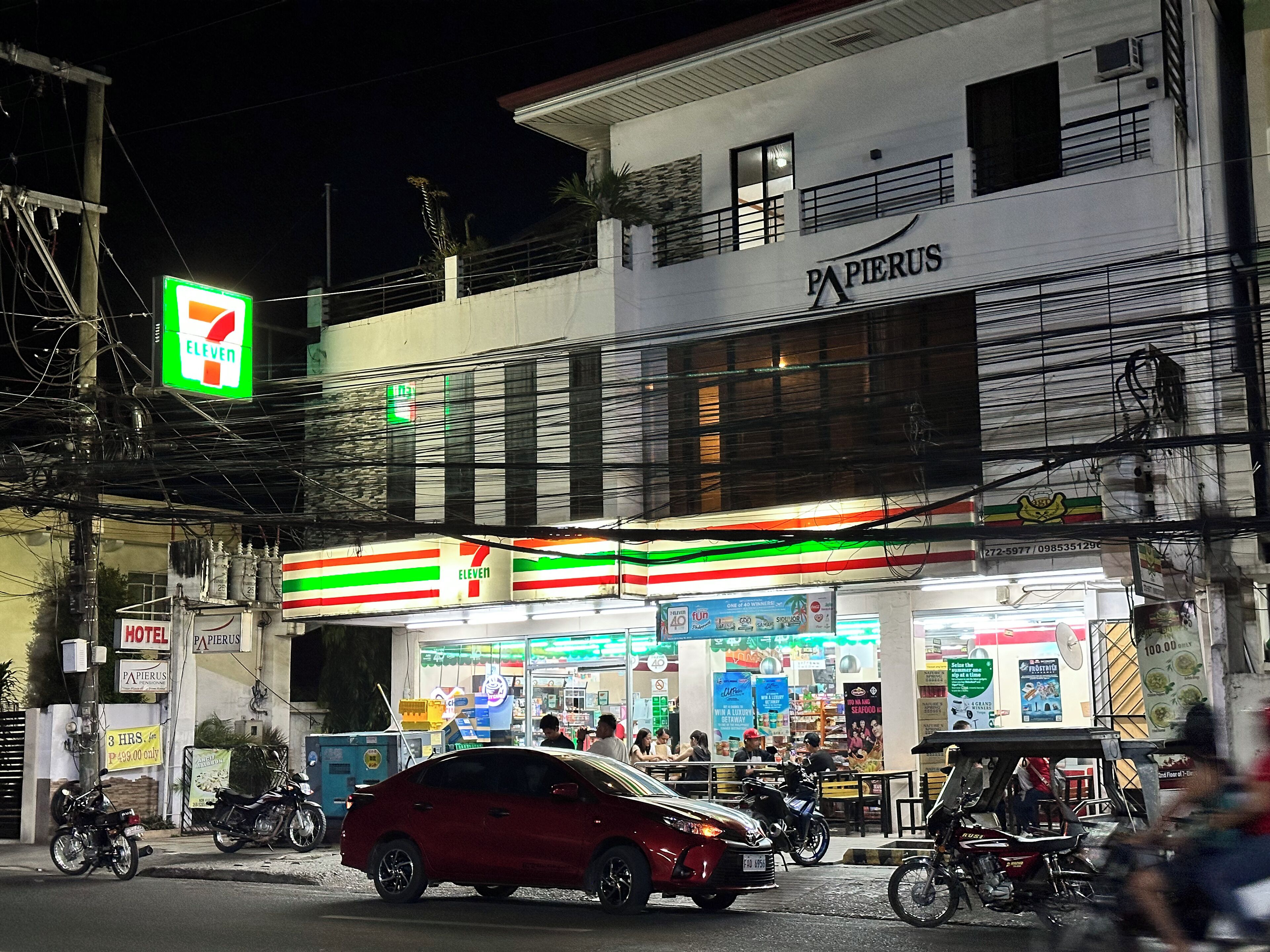 Uma loja de conveniência 7-Eleven com sinalização visível, ao lado de um edifício com a placa 'PAPIERUS'.