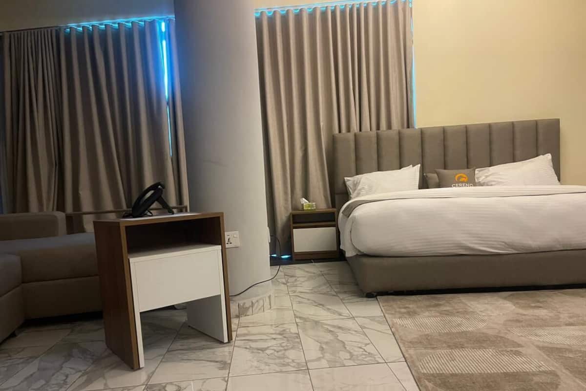 Quarto de hotel com cama, mesa de cabeceira, escrivaninha, cadeira e uma grande janela com cortinas.
