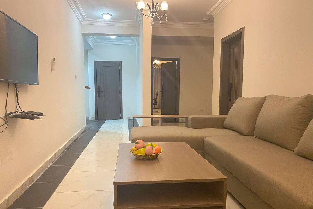 Uma sala de estar moderna com um sofá, uma mesa de centro com frutas, uma TV fixada na parede e um candelabro.