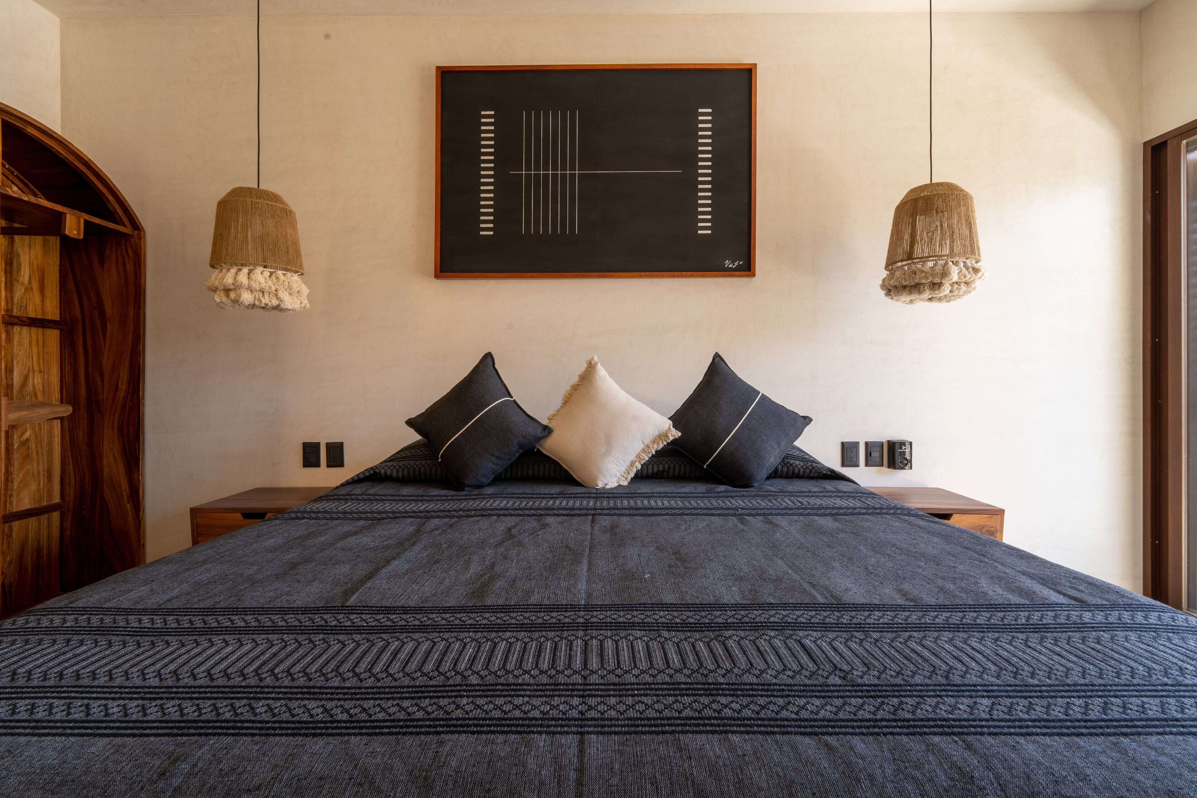 Quarto com uma cama grande, duas luminárias pendentes, uma obra de arte emoldurada na parede e uma porta de madeira.