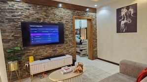 Uma sala de estar moderna com parede de pedra, TV de tela plana, mesa de centro de madeira e sofá cinza com almofadas vermelhas e pretas.
