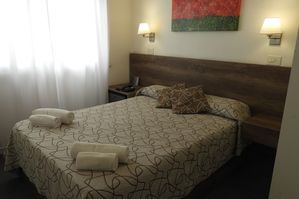 Quarto de hotel com uma cama, duas luminárias de cabeceira, um quadro na parede e uma mesinha de cabeceira com um telefone.