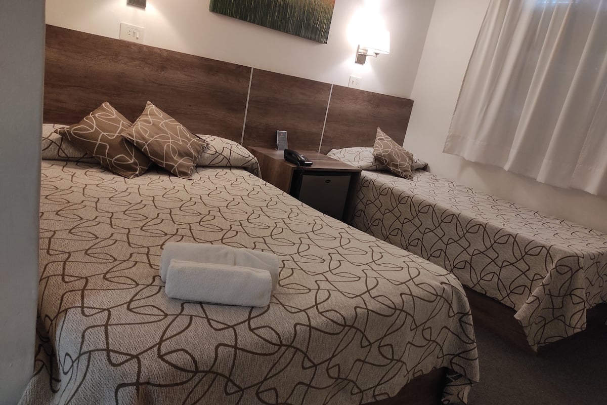 Quarto de hotel com duas camas, cada uma com roupa de cama estampada e cabeceira. Há uma mesa de cabeceira com um telefone e uma luminária.