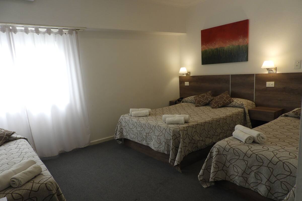 Quarto de hotel com duas camas de solteiro, uma janela grande com cortinas translúcidas, um quadro na parede e uma mesa de cabeceira com luminária.