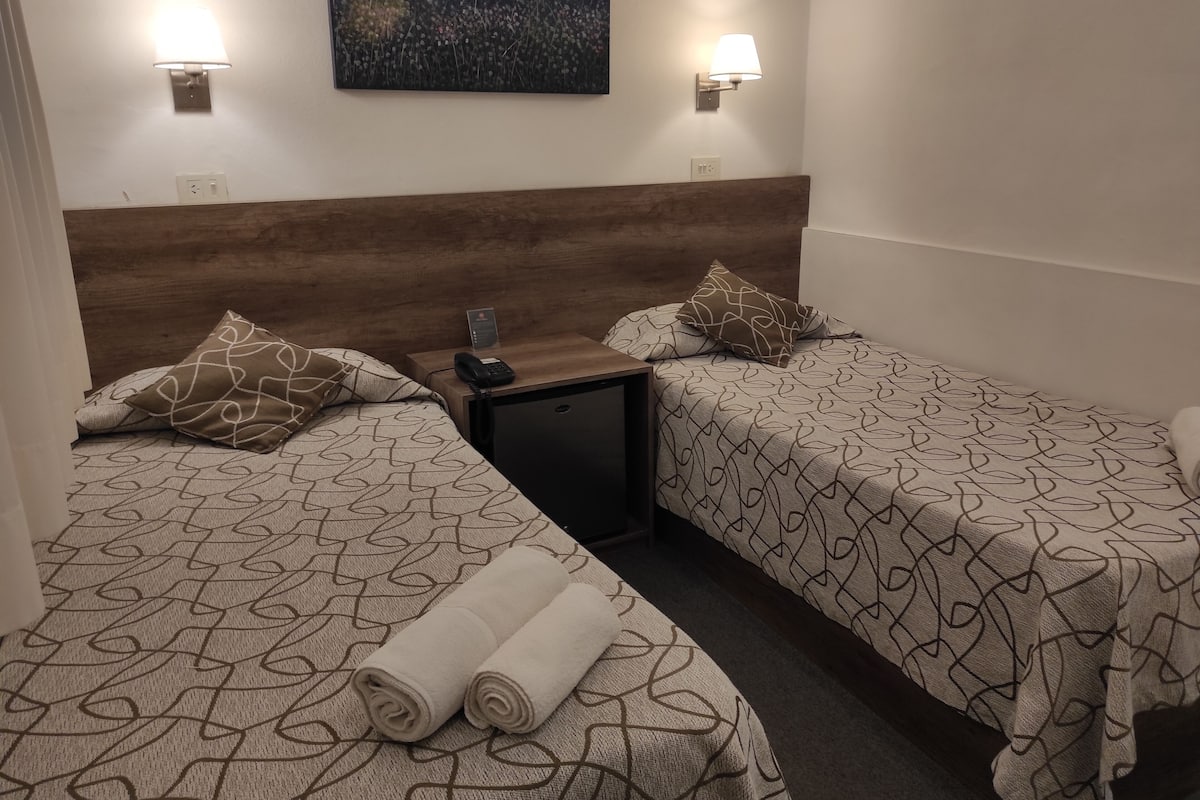 Quarto de hotel com duas camas, uma mesa de cabeceira, um telefone e um quadro na parede.