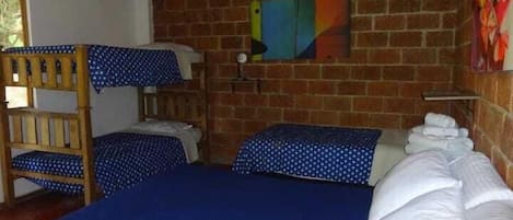 Quarto com parede de tijolos, duas beliches com roupa de cama azul, uma cadeira de madeira e uma prateleira com toalhas.