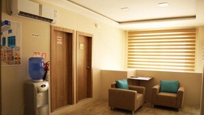 Quarto de hotel com duas portas de madeira, um dispensador de água, uma mesa pequena, dois poltronas e uma janela com persianas.