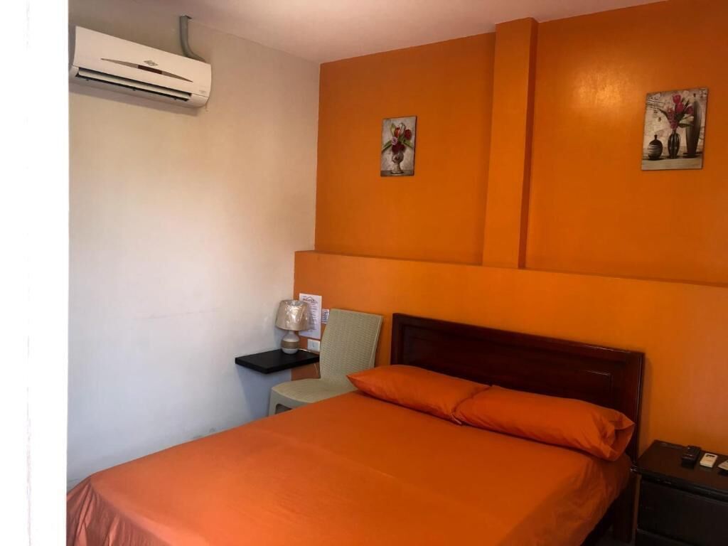 Quarto de hotel com cama, ar-condicionado, mesa de cabeceira, luminária e duas pinturas penduradas na parede.