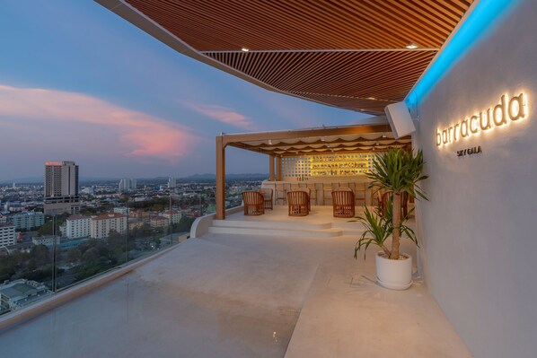 Um bar no terraço com vista para a cidade, um letreiro com a inscrição 'barracuda SKY GALA' e um vaso de planta.