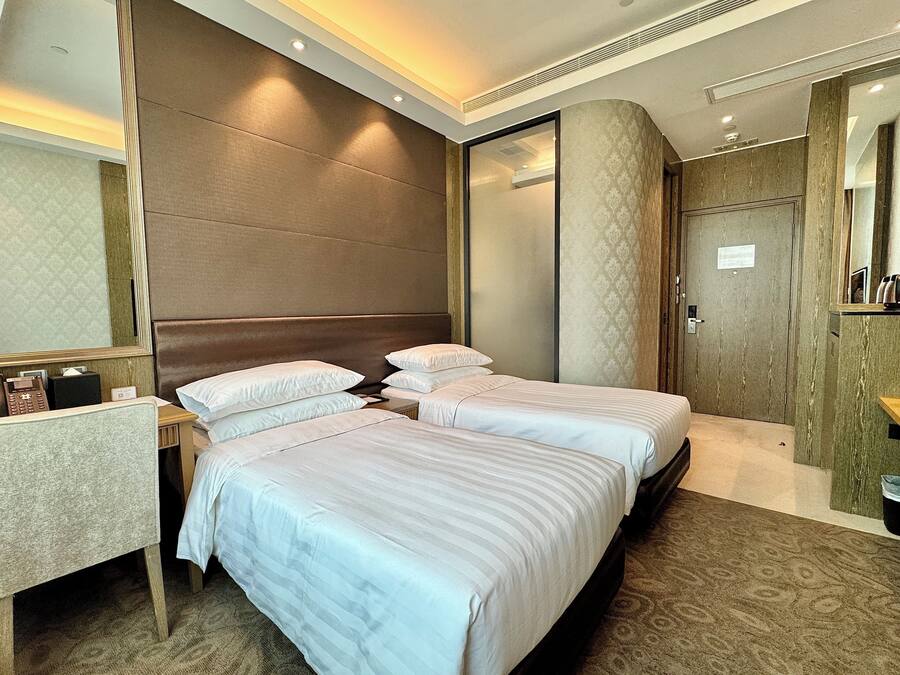 Quarto de hotel com duas camas, uma escrivaninha, um espelho e banheiro com chuveiro e vaso sanitário.