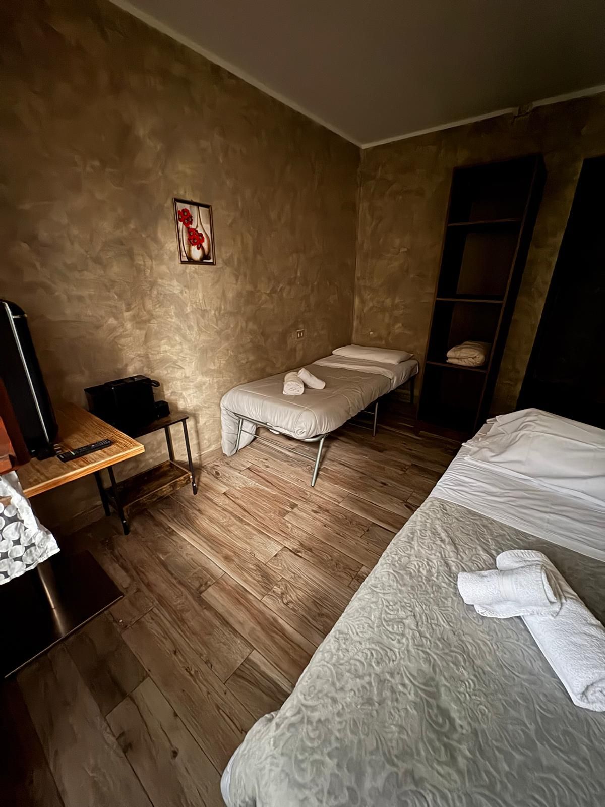 Quarto de hotel com piso de madeira, cama com colcha estampada, escrivaninha com computador e prateleira pequena com decoração de flor vermelha.