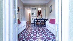 Um corredor com piso quadriculado, mobiliário branco e uma sala de jantar com cadeiras e mesa.