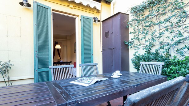 Uma mesa de madeira ao ar livre com cadeiras, um banco e uma porta com persianas verdes.