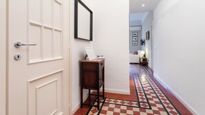 Um corredor com piso xadrez, uma porta com puxador, uma pequena mesa de madeira com uma placa e uma parede branca com um quadro emoldurado.