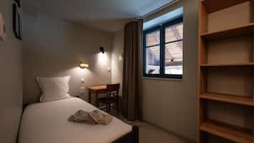 Quarto pequeno e bem organizado, com uma cama de solteiro, uma escrivaninha e uma janela com cortinas.