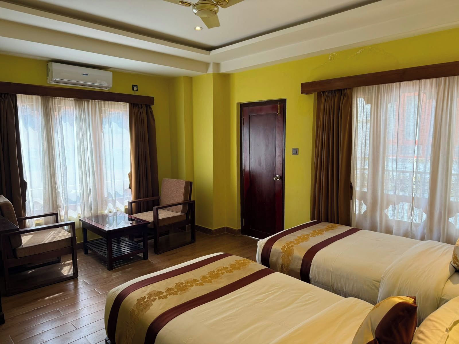 Quarto de hotel com duas camas, uma cadeira, uma mesinha e uma porta.