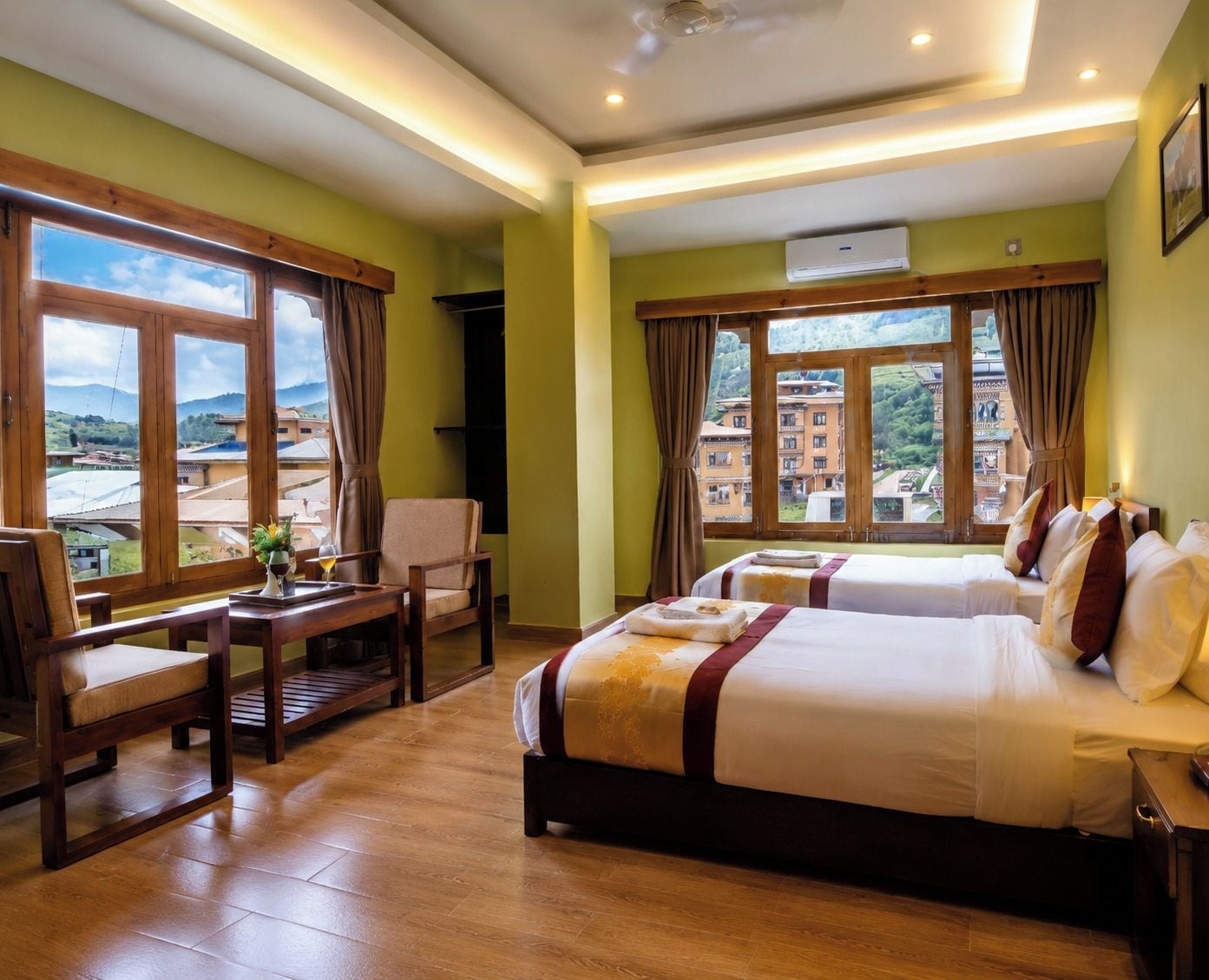 Quarto de hotel com cama, escrivaninha, cadeira e janelas amplas com vista para edifícios e montanhas.