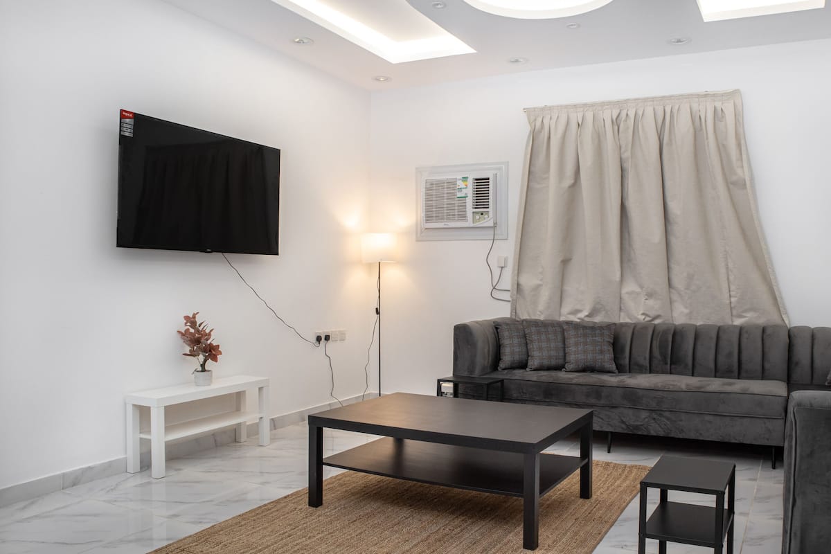 Uma sala de estar moderna com um sofá cinzento, uma mesa de centro preta, uma mesinha de cabeceira branca, uma TV de tela plana na parede e um aparelho de ar condicionado.