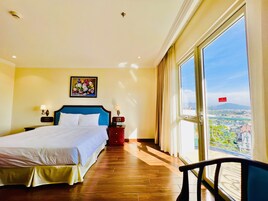 Quarto de hotel com uma cama grande, mesa de cabeceira com abajur, quadro na parede e varanda com vista.
