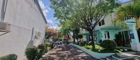 Rua residencial com calçada pavimentada, caminho arborizado e prédio com fachada branca e unidade de ar condicionado.