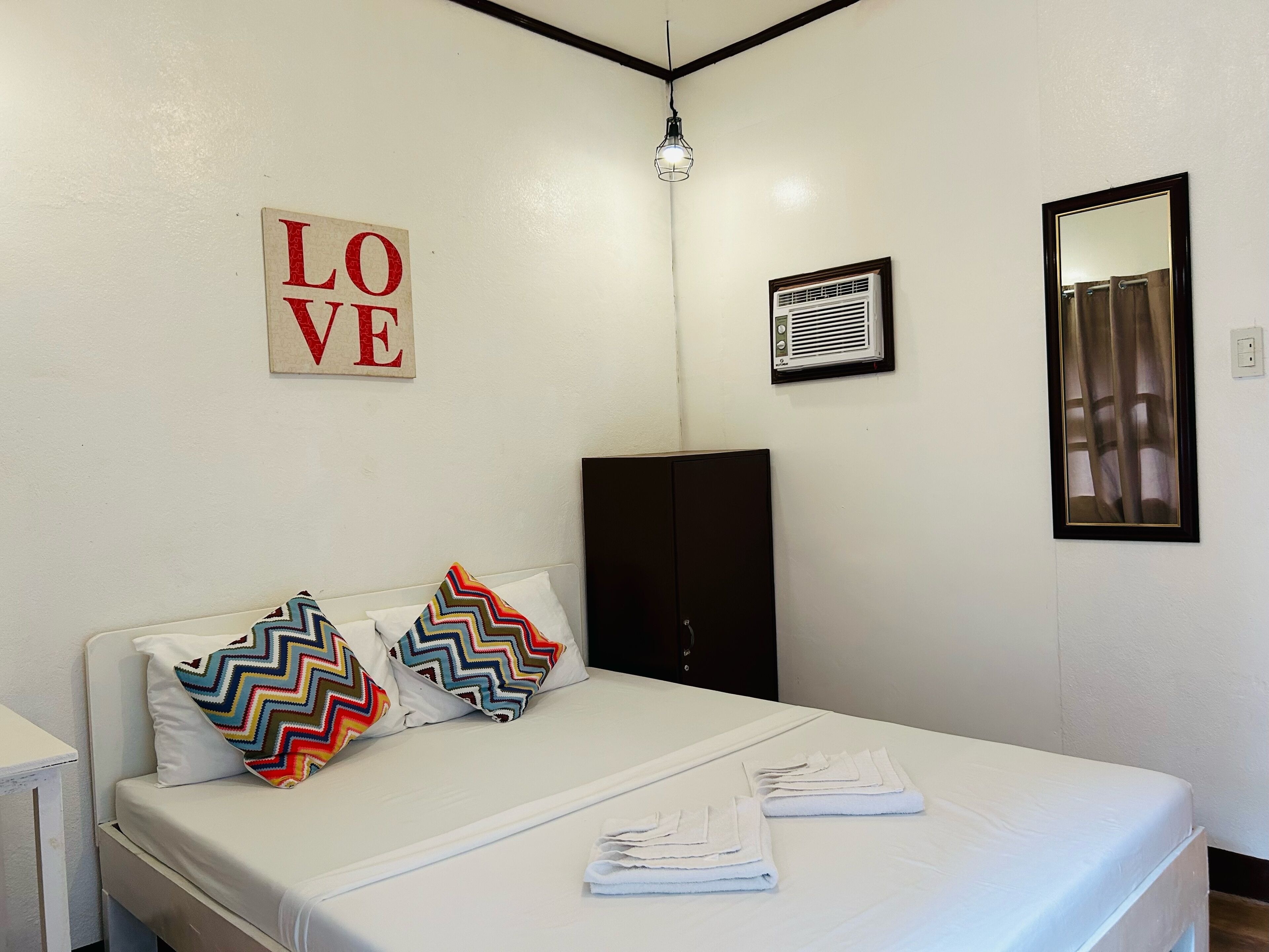 Cama bem arrumada com duas almofadas coloridas, ar condicionado fixado na parede, um espelho e um letreiro com a palavra 'LOVE'.