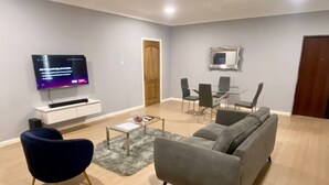 Uma sala de estar com um sofá cinzento, uma poltrona azul, uma mesa de centro de vidro, uma televisão sobre um suporte branco e uma área de jantar com mesa e cadeiras de vidro.